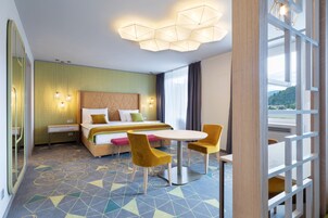 Quarto de hotel com uma cama, mesa de cabeceira, duas cadeiras, uma mesa redonda e uma janela grande com vista.
