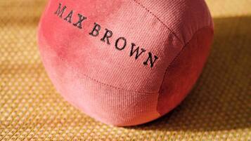 Um objeto redondo e rosa com a inscrição "MAX BROWN" bordada, sobre uma superfície de tecido trançado.
