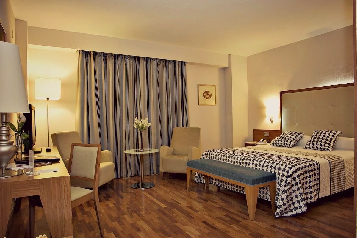 Quarto de hotel com cama, escrivaninha, cadeira, mesa pequena com um vaso e uma janela grande com cortinas.