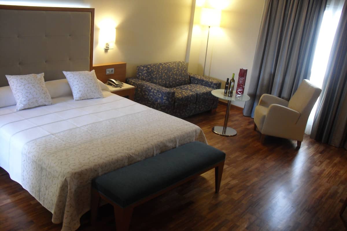 Quarto de hotel com uma cama grande, um banco, uma cadeira, um sofá, uma mesinha e um abajur.