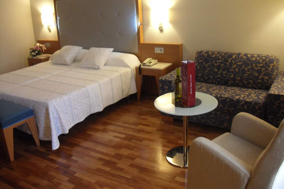 Quarto de hotel com cama, sofá, uma mesinha e uma cadeira. Há uma mesinha de cabeceira com abajur e telefone.