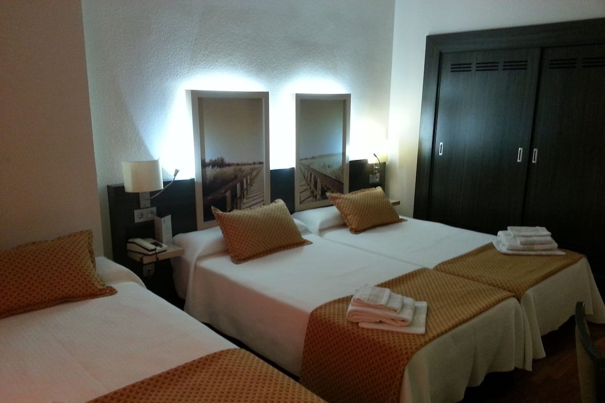 Quarto de hotel com duas camas de solteiro, uma escrivaninha com abajur, um guarda-roupa e um quadro na parede.