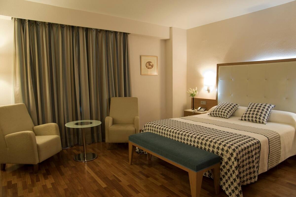 Quarto de hotel com uma cama grande, duas poltronas, uma mesa redonda pequena e um banquinho.