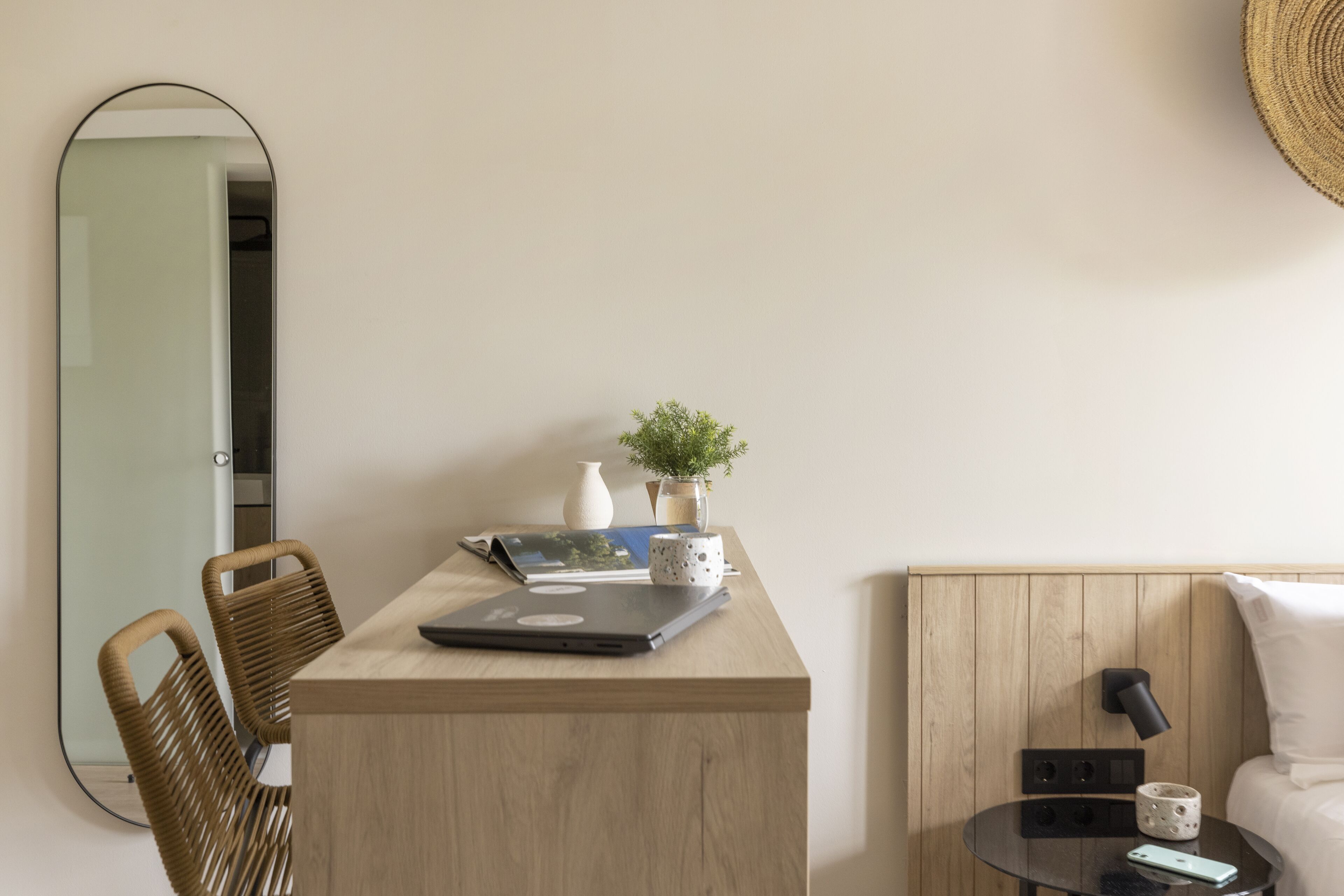 Quarto moderno com escrivaninha de madeira, um laptop, uma pequena planta e mesa de cabeceira com abajur e telefone.