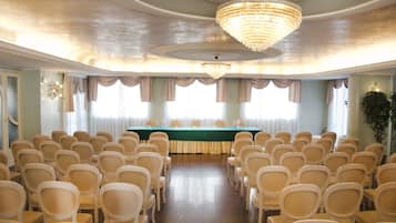 Sala de conferências com fileiras de cadeiras voltadas para uma mesa, um candelabro e grandes janelas com cortinas.