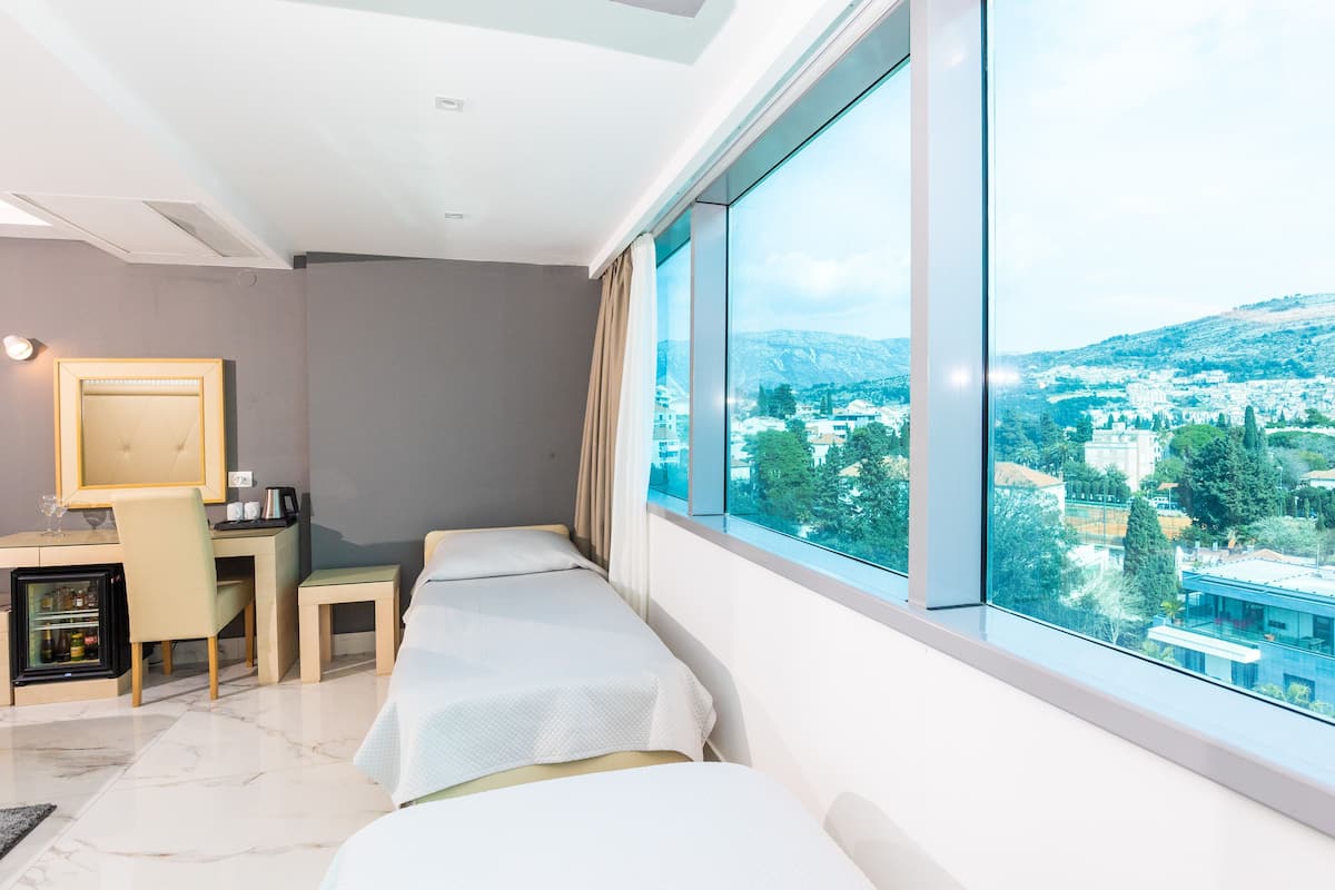 Quarto de hotel moderno com uma janela grande, uma escrivaninha com cadeira, um minibar e duas camas.