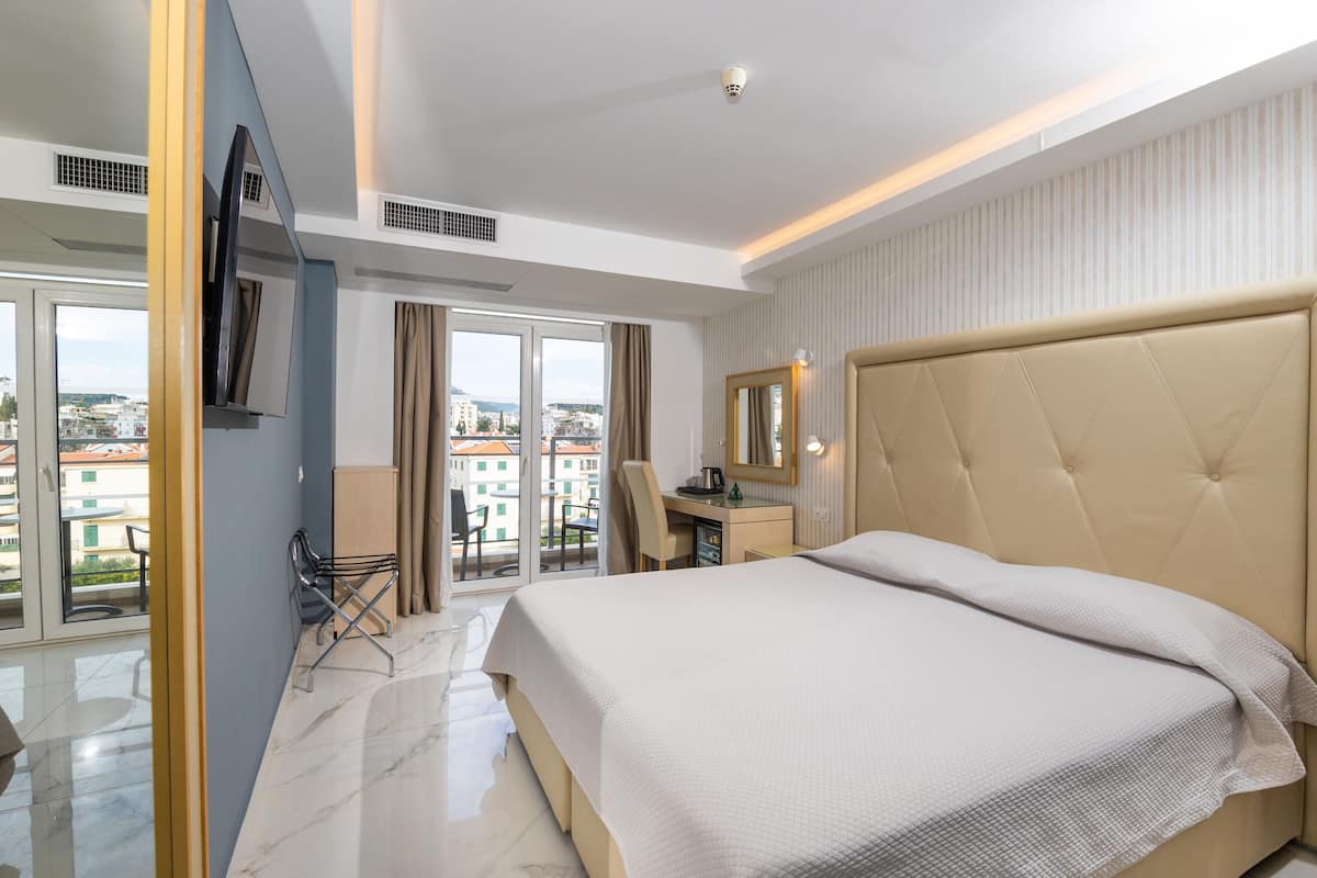 Quarto de hotel com cama grande, escrivaninha, cadeira e varanda com vista para a cidade.