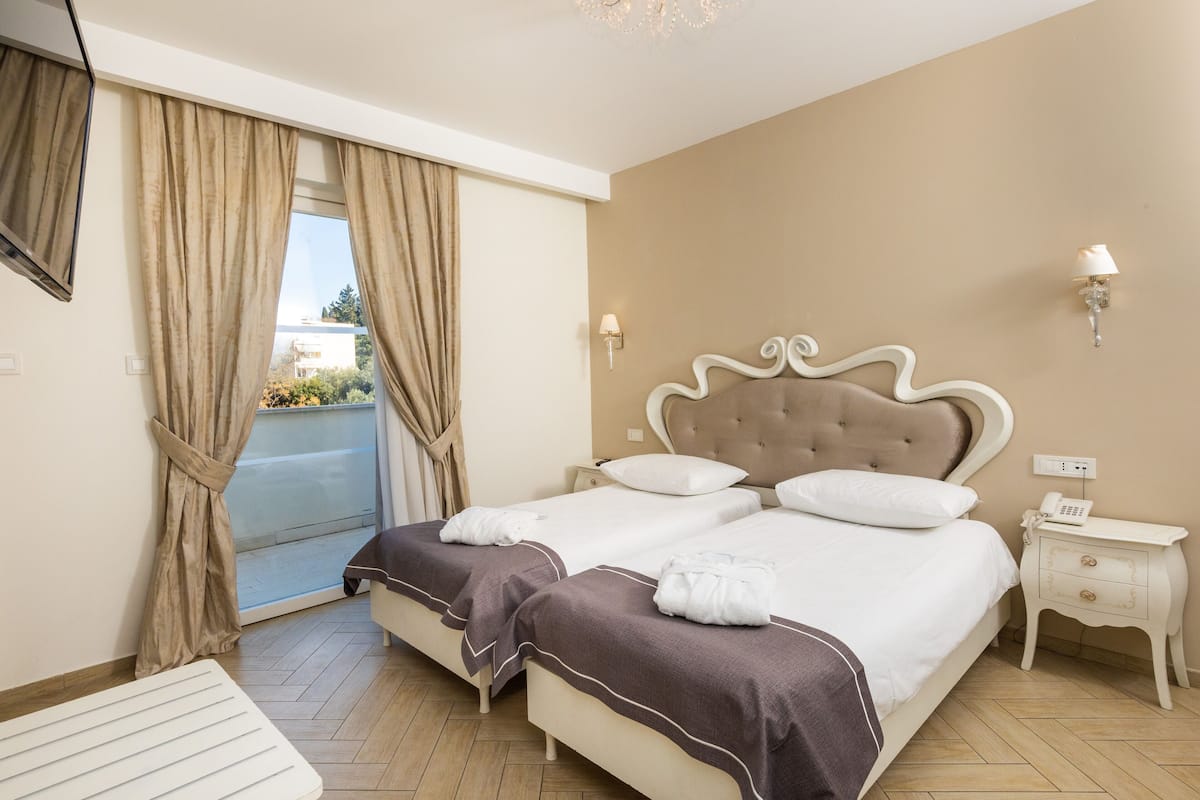 Uma cama de casal com cabeceira acolchoada, duas mesas de cabeceira com luminárias, vista através de uma porta de vidro e uma pequena mesa com uma luminária.