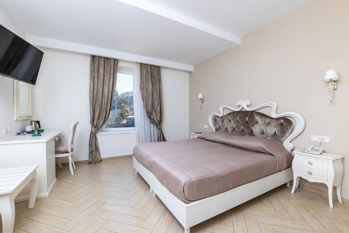 Quarto com cama, escrivaninha, cadeira, televisão e janela com cortinas.