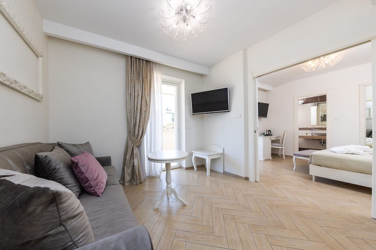 Uma sala de estar moderna com um sofá, uma mesinha redonda, uma televisão na parede e um quarto com uma cama e uma escrivaninha.