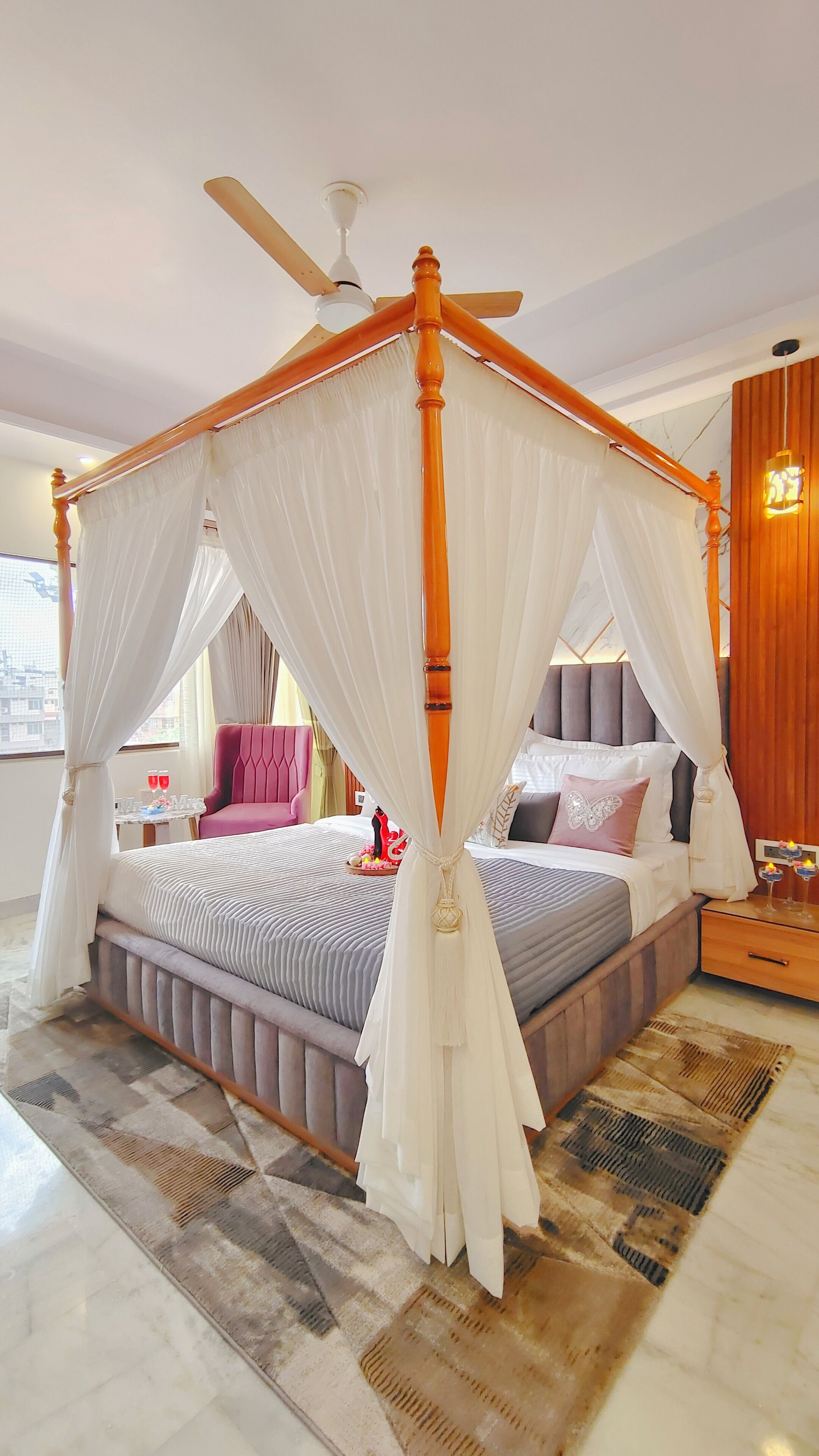 Uma cama com dossel e cortina, um ventilador no teto, uma mesinha de cabeceira com abajur e vista para a cidade pela janela.