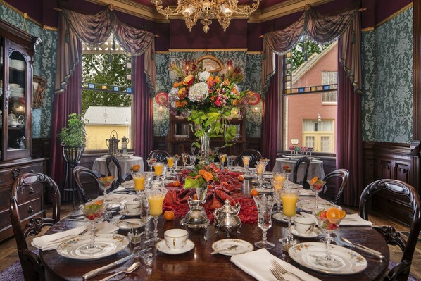 Uma sala de jantar formal com uma mesa grande posta para refeição, rodeada por cadeiras e um arranjo decorativo.