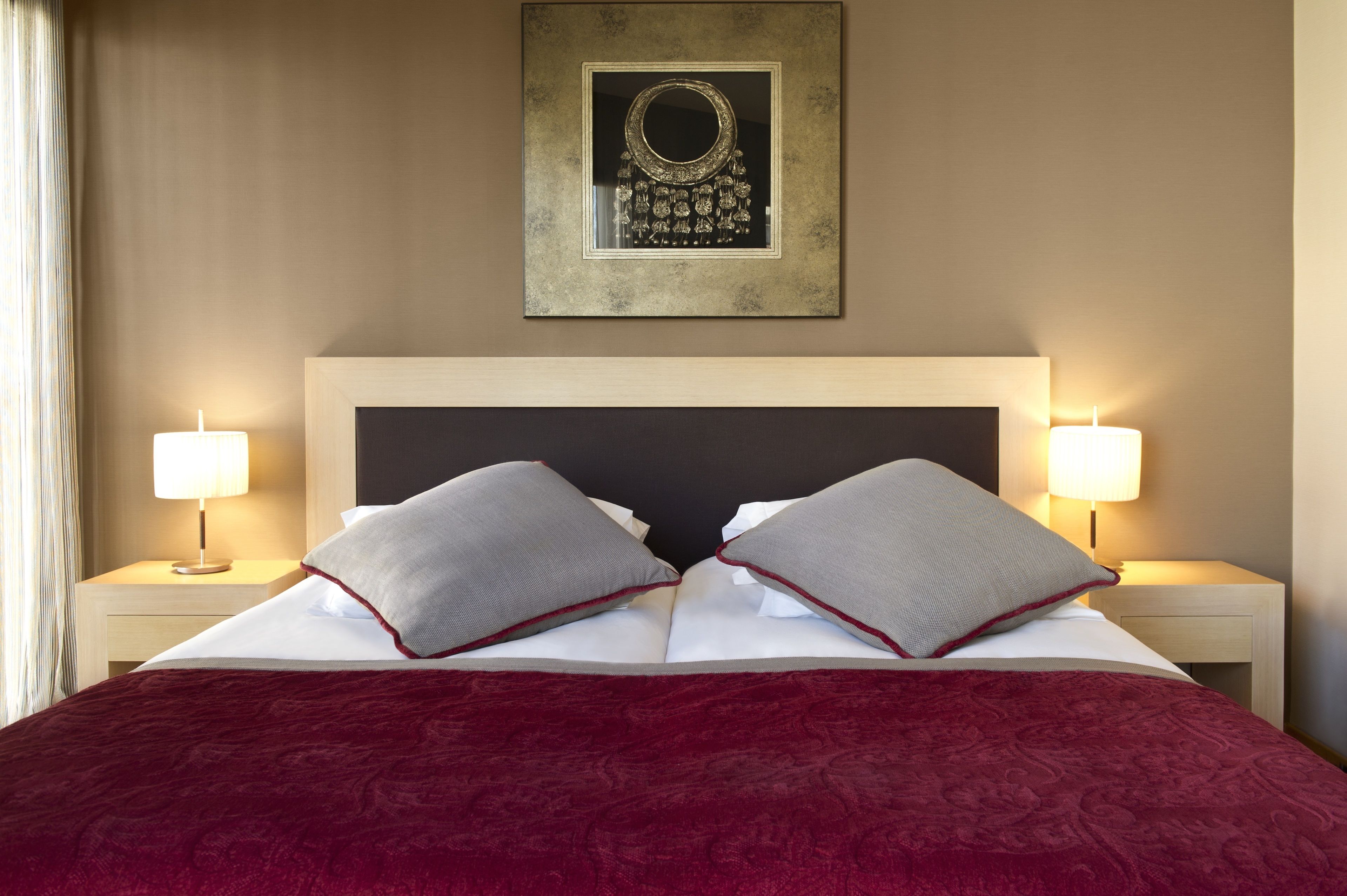 Cama bem arrumada com um cobertor vermelho, duas almofadas e uma obra de arte emoldurada acima. Duas luminárias de cabeceira em ambos os lados da cama.