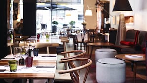 Interior moderno de restaurante com mesas e cadeiras de madeira, e opções de assento variadas, incluindo um sofá e um banquinho.