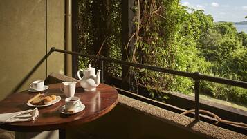 Uma mesa de madeira com um bule branco, xícaras e doces numa varanda com vista para uma paisagem verde exuberante e um corpo d'água.