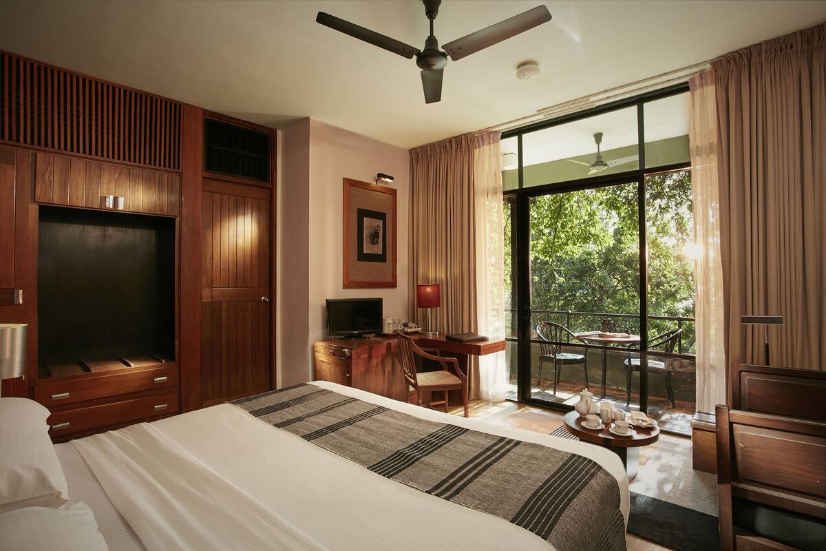 Quarto de hotel com uma cama grande, uma escrivaninha com computador, televisão e varanda com mesa e cadeiras.