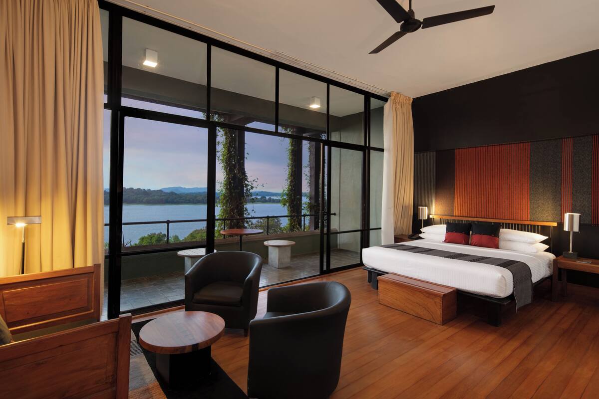Quarto de hotel moderno com uma cama grande, duas cadeiras, uma mesinha e varanda com vista para a água.
