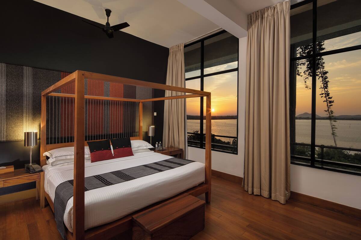 Quarto moderno com cama de dossel, uma janela grande com cortinas e vista para o pôr do sol sobre o mar.