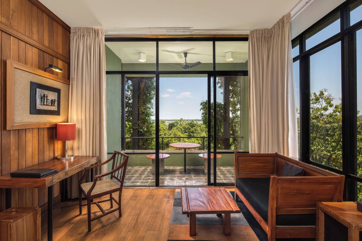 Quarto com escrivaninha de madeira, cadeira, sofá, uma pequena mesa e vista através de grandes janelas.