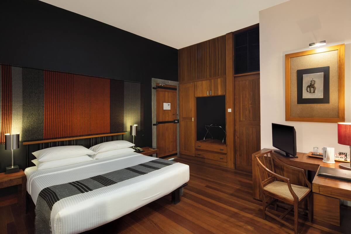 Quarto de hotel com cama grande, guarda-roupa de madeira, escrivaninha, cadeira e um quadro na parede.