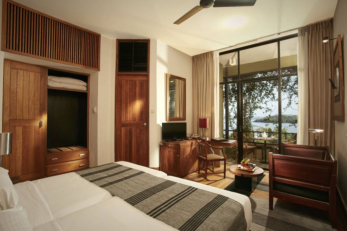 Quarto de hotel com uma cama grande, guarda-roupa de madeira, escrivaninha, cadeira e varanda com vista.