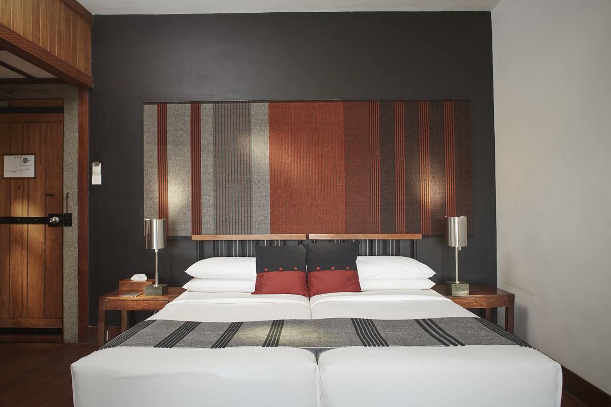 Quarto de hotel com uma cama grande, duas mesas de cabeceira, uma porta de madeira e cabeceira escura com listras verticais.