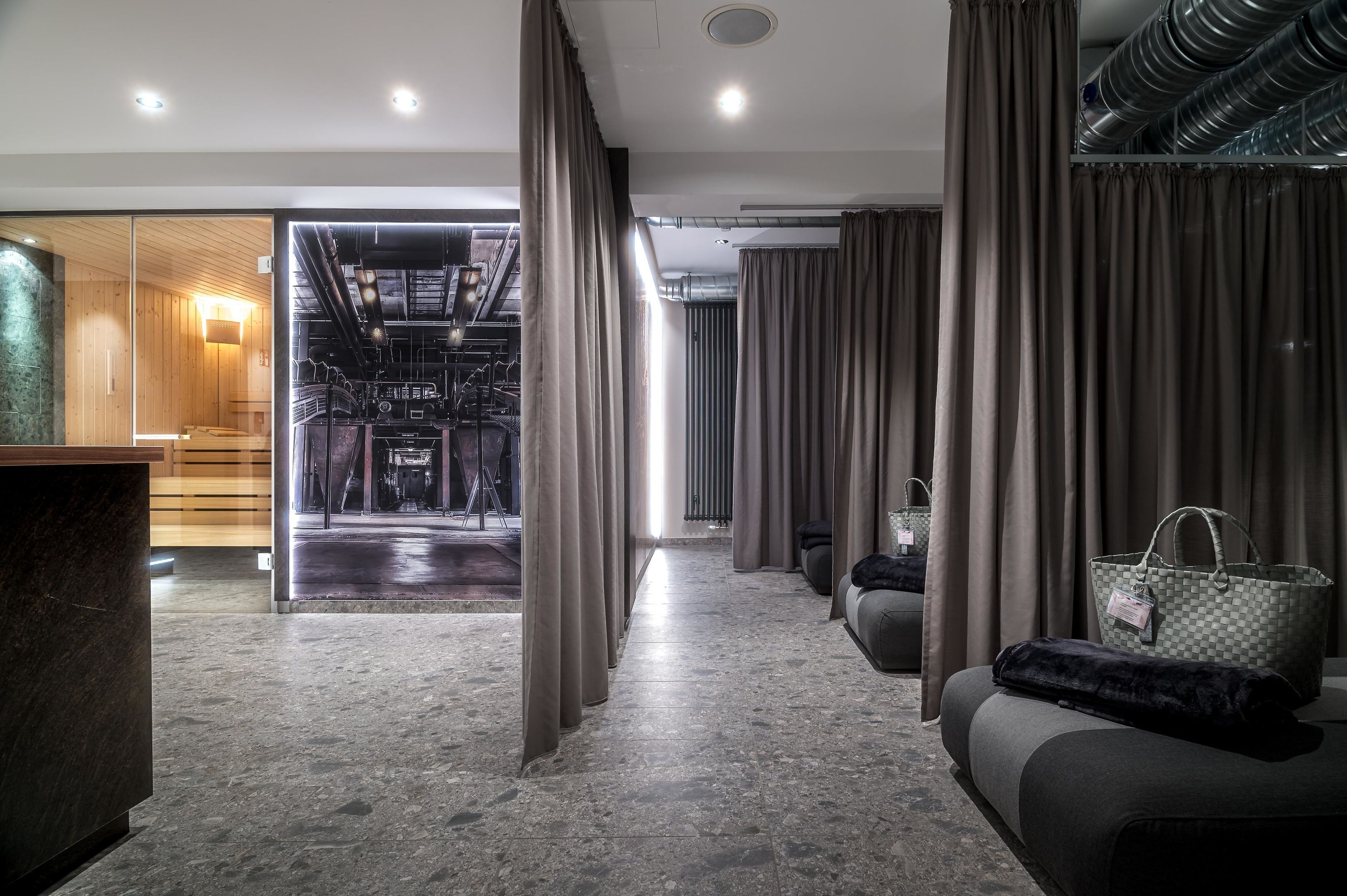 Um lobby de hotel moderno com uma receção, área de estar com um sofá e uma mala verde quadriculada grande, e vista para um quarto com soalho de madeira e uma escadaria.
