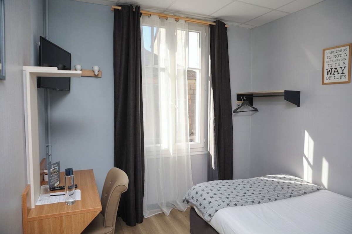 Quarto de hotel com uma cama, uma escrivaninha, uma cadeira, uma televisão e uma janela com cortinas.