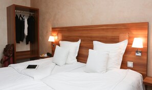 Quarto de hotel com cabeceira de madeira, roupa de cama branca, mesa de cabeceira com abajur e um cabideiro com roupas.
