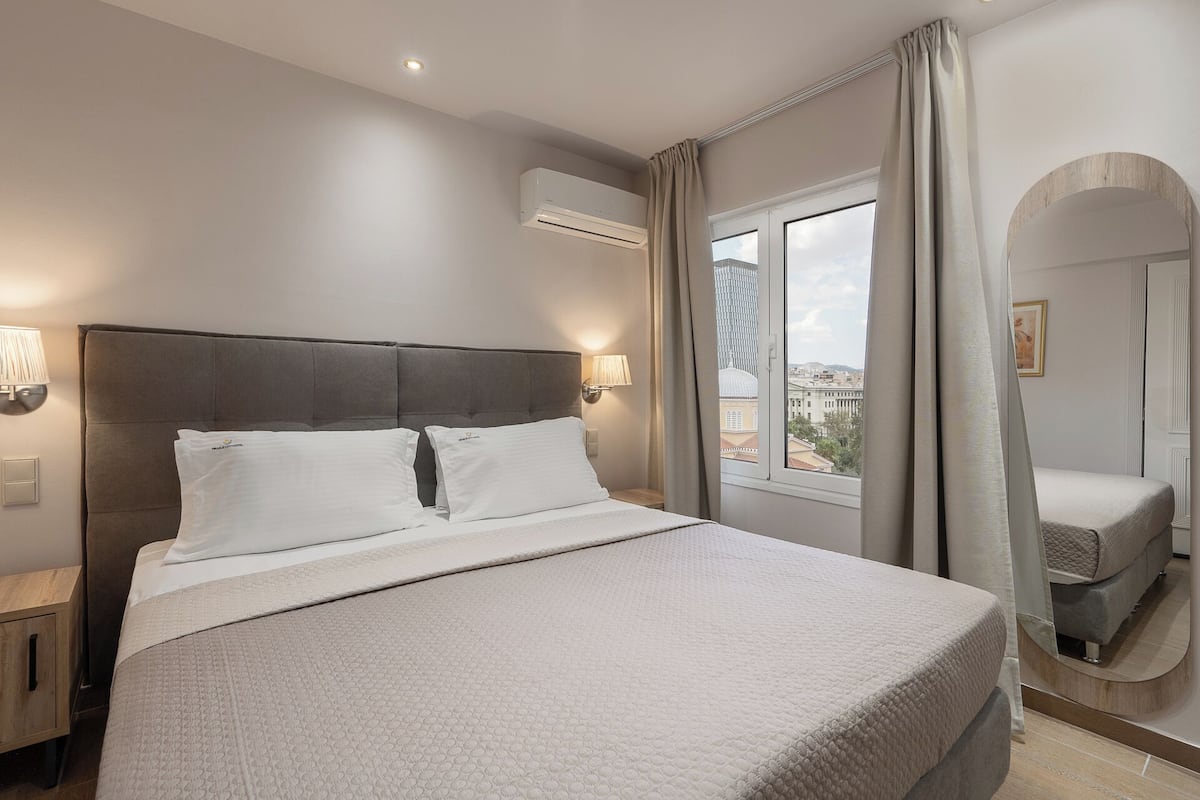 Quarto de hotel moderno com uma cama grande, cabeceira, luminária de cabeceira, aparelho de ar condicionado e vista para a cidade pela janela.