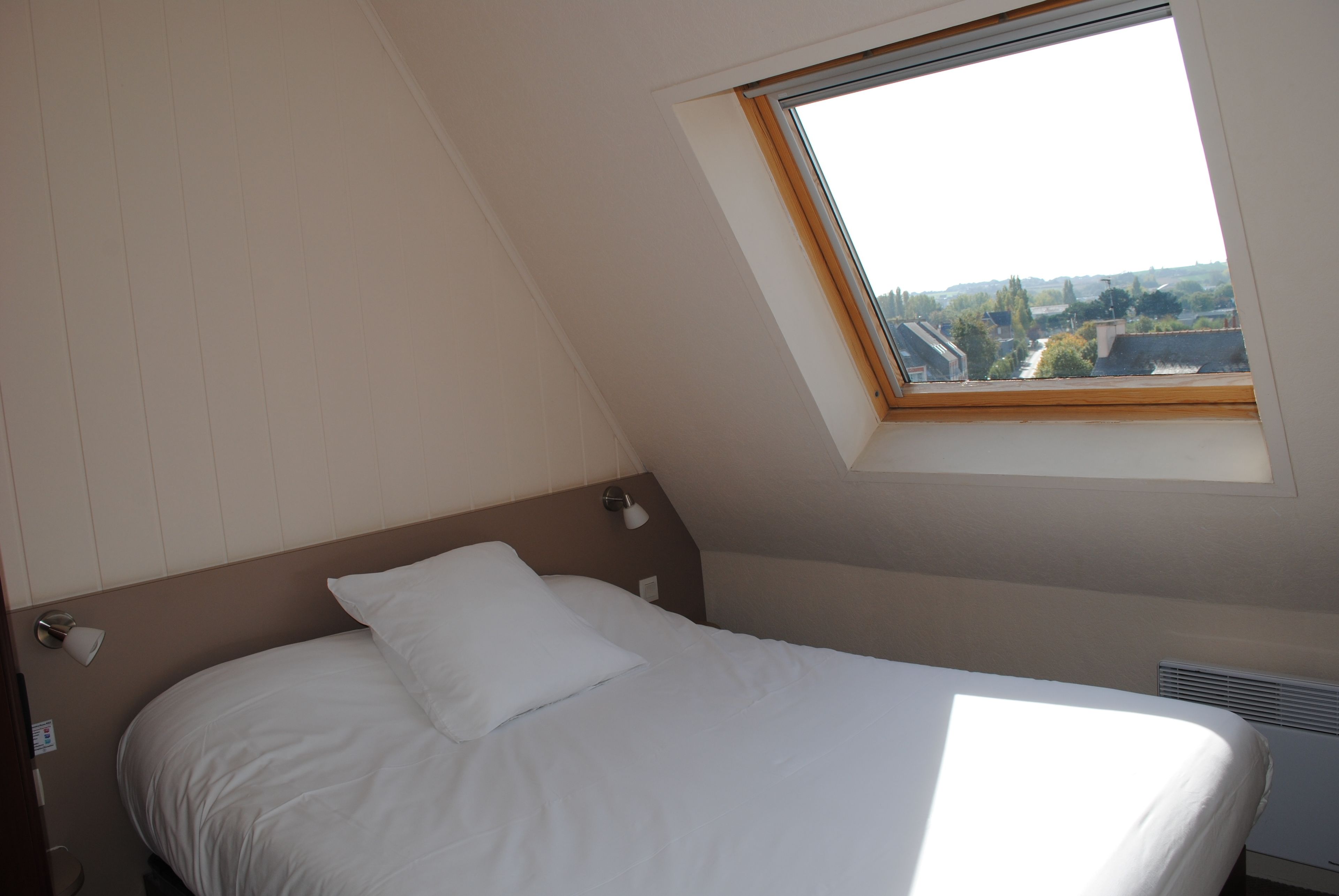Quarto com teto inclinado, uma cama de solteiro, janela com vista para a paisagem urbana e luminária fixada na parede.