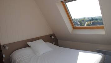Quarto com teto inclinado, uma cama de solteiro, janela com vista para a paisagem urbana e luminária fixada na parede.