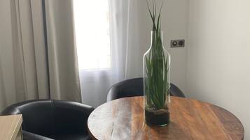 Um quarto com uma mesa redonda de madeira, duas cadeiras de couro preto, um vaso de vidro com plantas verdes, uma janela com cortinas brancas e uma tomada de parede.