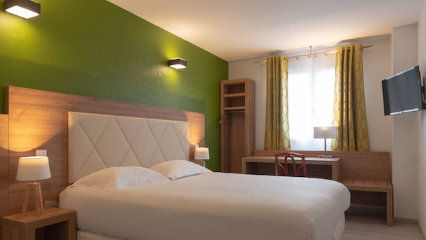 Quarto de hotel com uma cama grande, luminárias de cabeceira, uma escrivaninha, uma cadeira, uma televisão e um guarda-roupa.