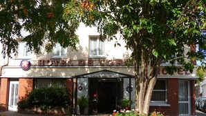 Entrada de hotel com fachada de tijolos, uma grande árvore na frente e um canteiro de flores.
