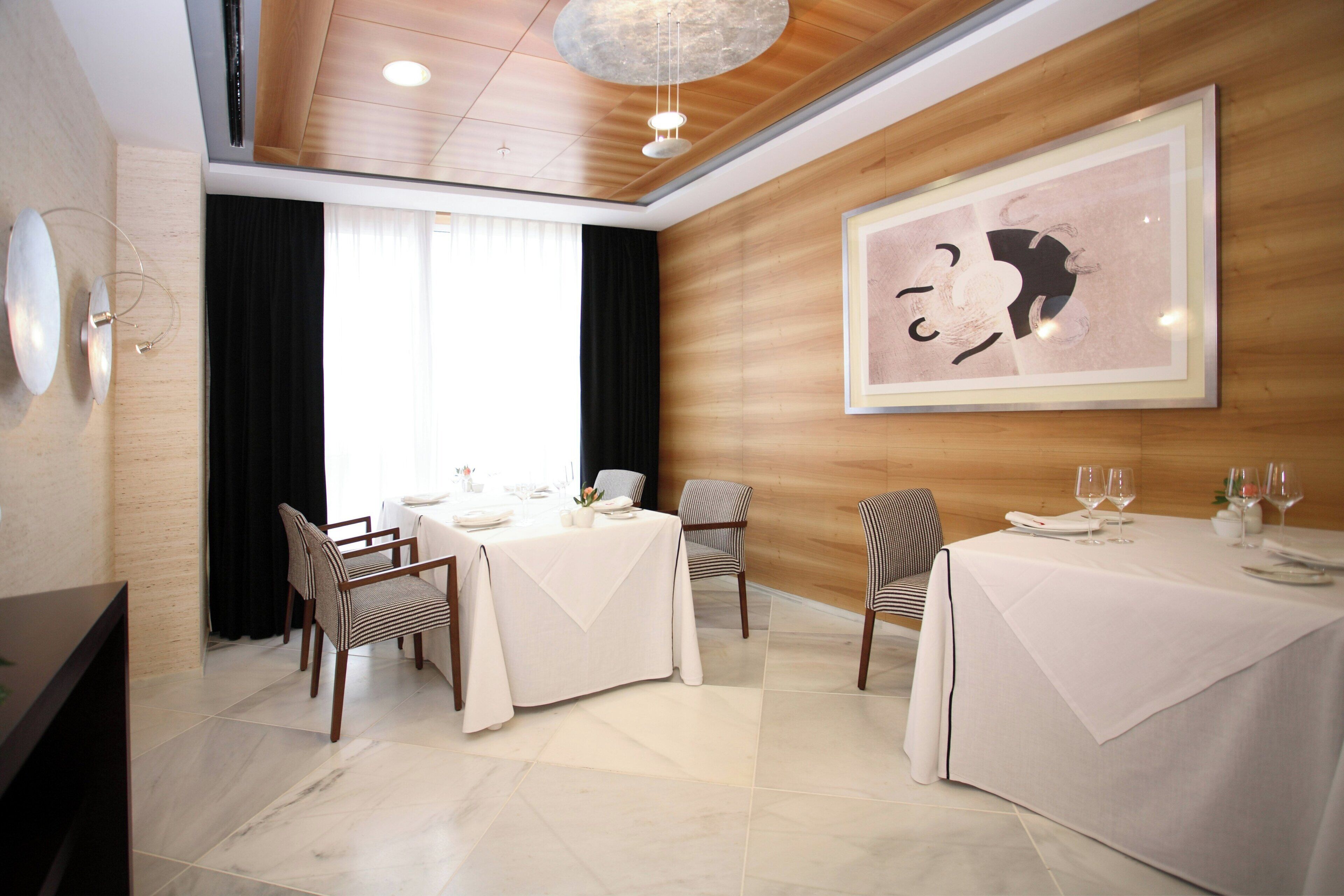 Uma sala de jantar com duas mesas, cadeiras, um lustre e uma obra de arte emoldurada na parede.