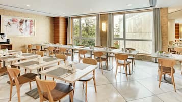 Uma área de refeições moderna com várias mesas e cadeiras, grandes janelas com vista para árvores e uma máquina de café fixada na parede.