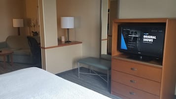 Quarto de hotel com uma cama, uma escrivaninha com uma televisão exibindo 'ORIGINAL SHOWS', uma cadeira, um abajur e uma pequena mesa.
