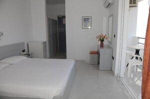 Quarto de hotel com cama, escrivaninha com cadeira, mesinha com um vaso de flores e porta que dá para outro cômodo.