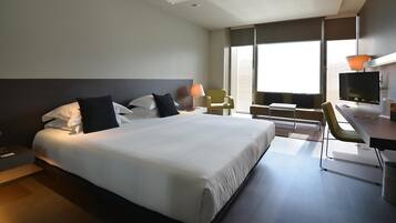 Quarto de hotel com uma cama grande, uma escrivaninha com cadeira, televisão e janela com persianas.