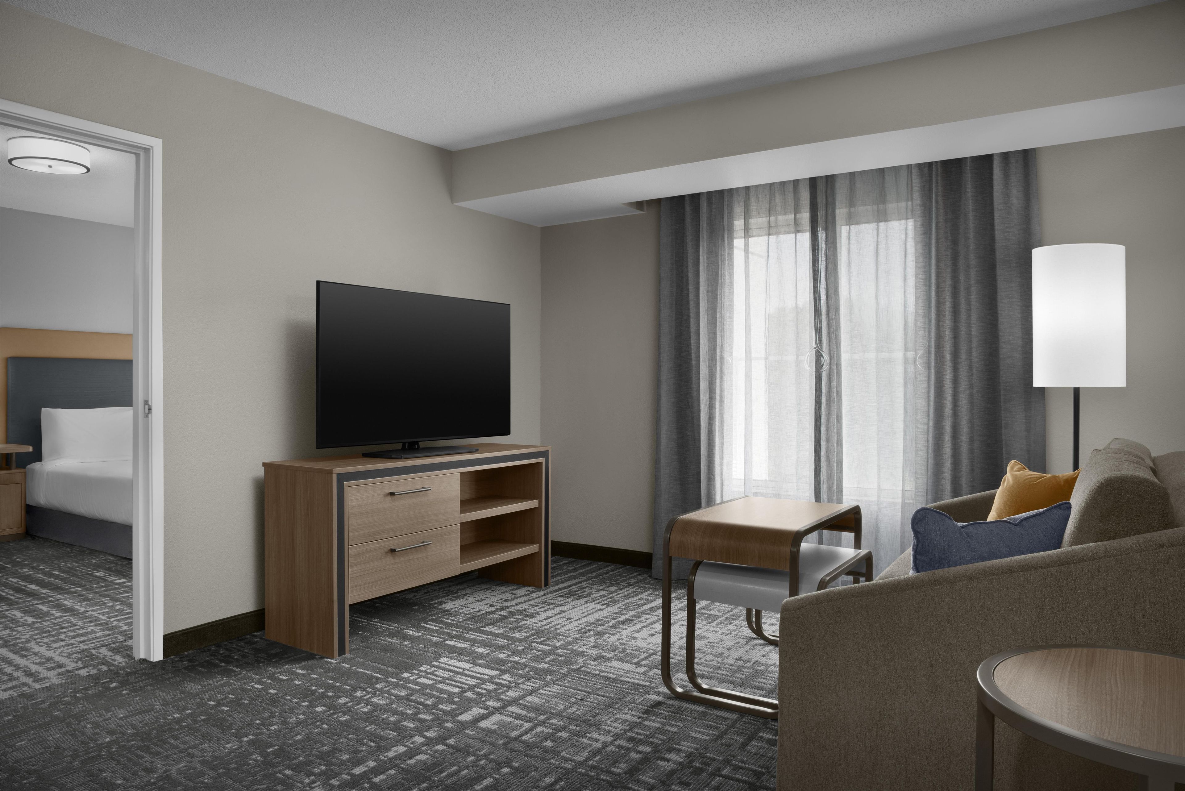 Quarto de hotel com televisão de tela plana em suporte de madeira, escrivaninha com cadeira, sofá e uma janela grande com cortinas.