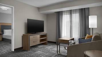 Quarto de hotel com televisão de tela plana em suporte de madeira, escrivaninha com cadeira, sofá e uma janela grande com cortinas.