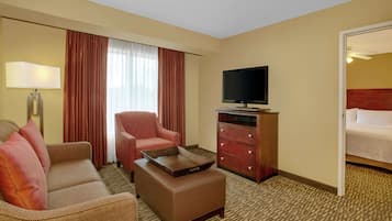Quarto de hotel com sofá, poltrona, banquinho, televisão, cômoda e cama.