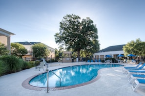 Piscina com espreguiçadeiras e guarda-sóis, cercada por vegetação e edifícios.