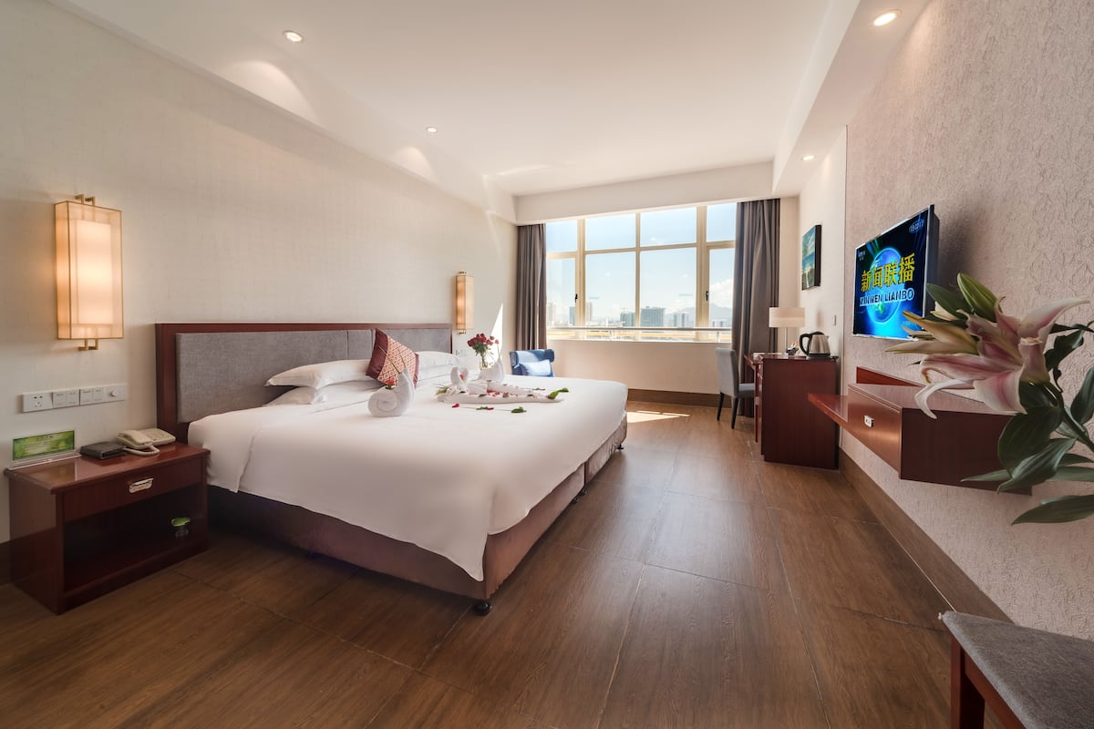 Quarto de hotel com cama grande, mesa de cabeceira, luminária, TV e vista para a cidade pela janela.