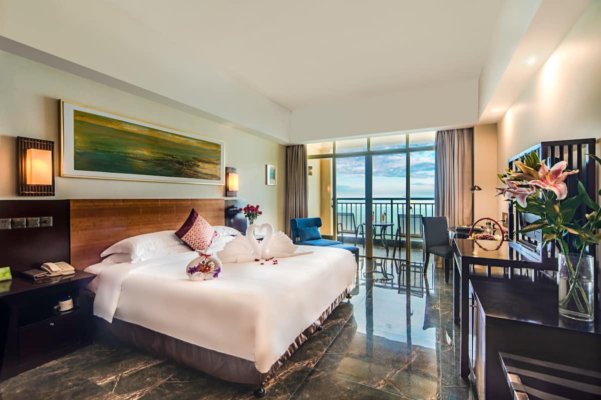 Quarto de hotel com cama grande, escrivaninha, cadeira e varanda com vista para o oceano.