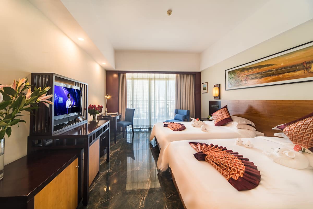 Quarto de hotel com duas camas, uma escrivaninha com televisão, uma janela grande com cortinas e um quadro na parede.