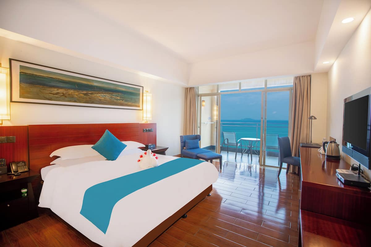 Quarto de hotel com uma cama grande, uma mesa com televisão, uma varanda com vista e um quadro na parede.
