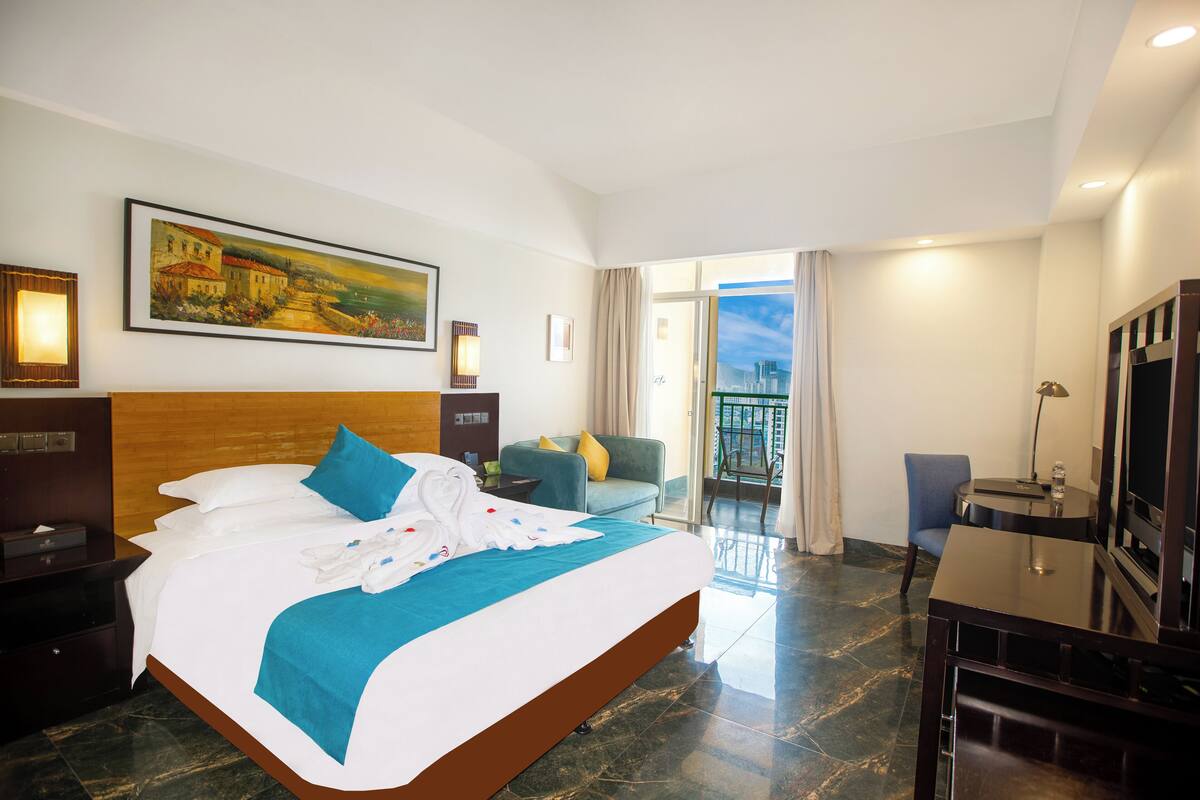 Quarto de hotel com uma cama grande, uma escrivaninha, uma cadeira, uma televisão e uma varanda com vista para a cidade.