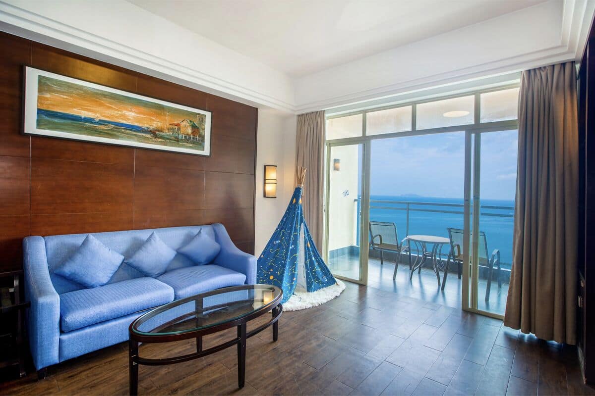 Quarto com sofá azul, mesa de centro de vidro, pintura na parede e vista para o mar através de grandes janelas.